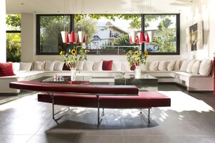 Uma sala de estar moderna com uma janela grande, uma mesa de centro de vidro, um banco vermelho e um sofá com almofadas brancas.