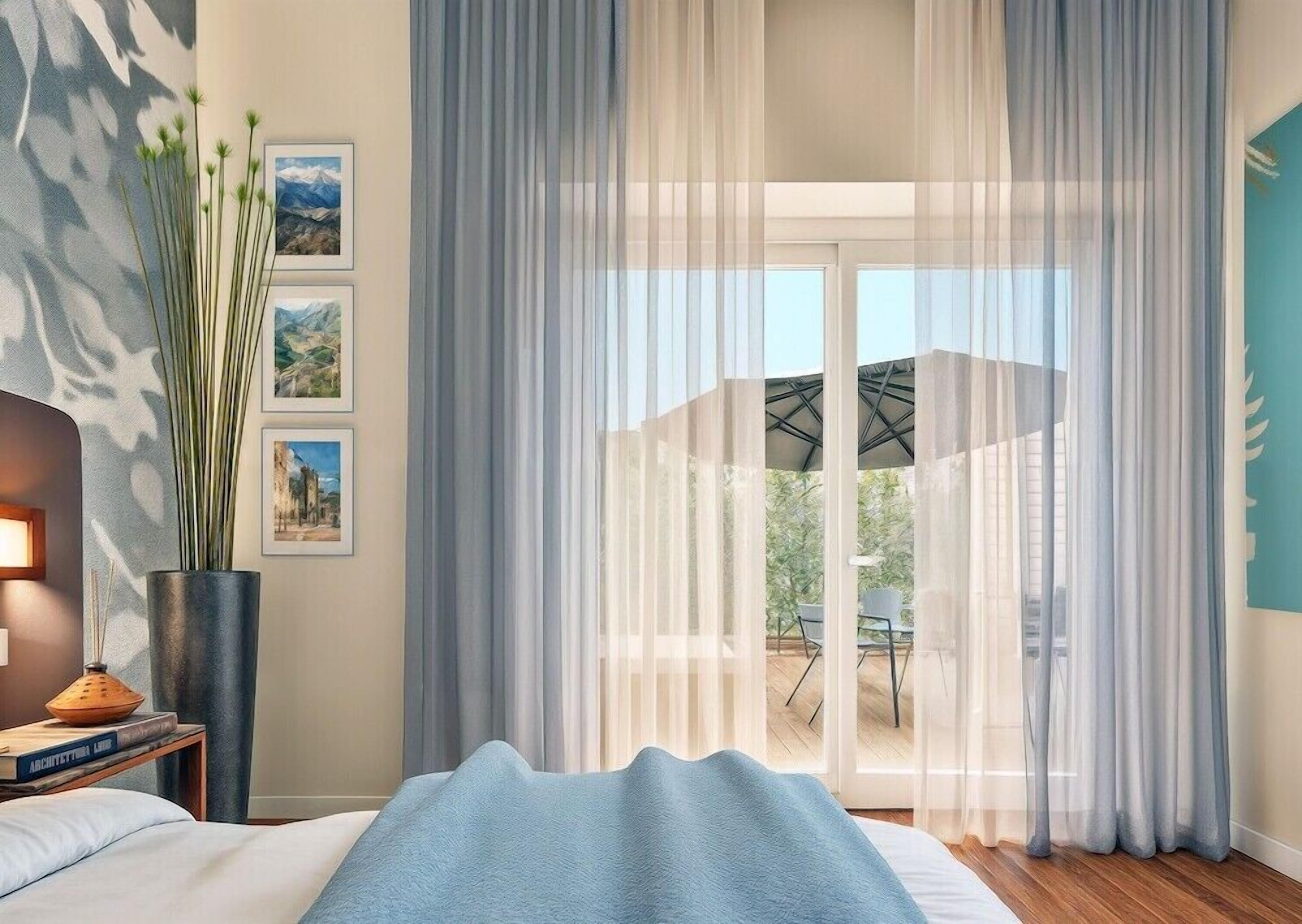 Quarto com cama, mesa de cabeceira, um vaso com plantas e vista para o exterior através de uma porta de vidro.