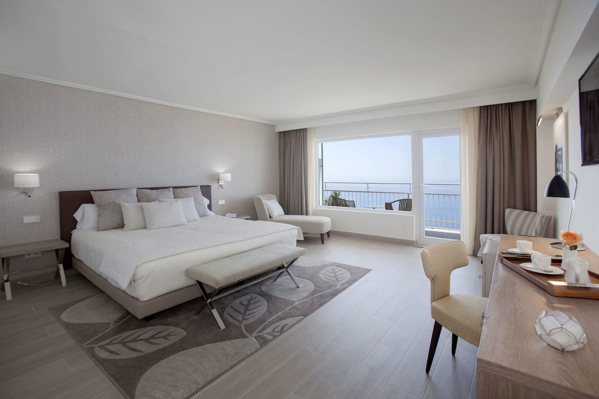 Quarto de hotel moderno com uma cama grande, uma escrivaninha com cadeira, um sofá pequeno e uma varanda com vista.