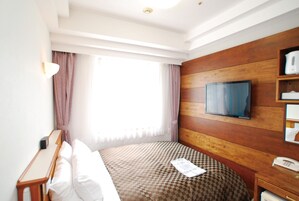 Quarto de hotel com uma cama grande, televisão fixada na parede de madeira, mesa de cabeceira com abajur e uma janela com cortinas.