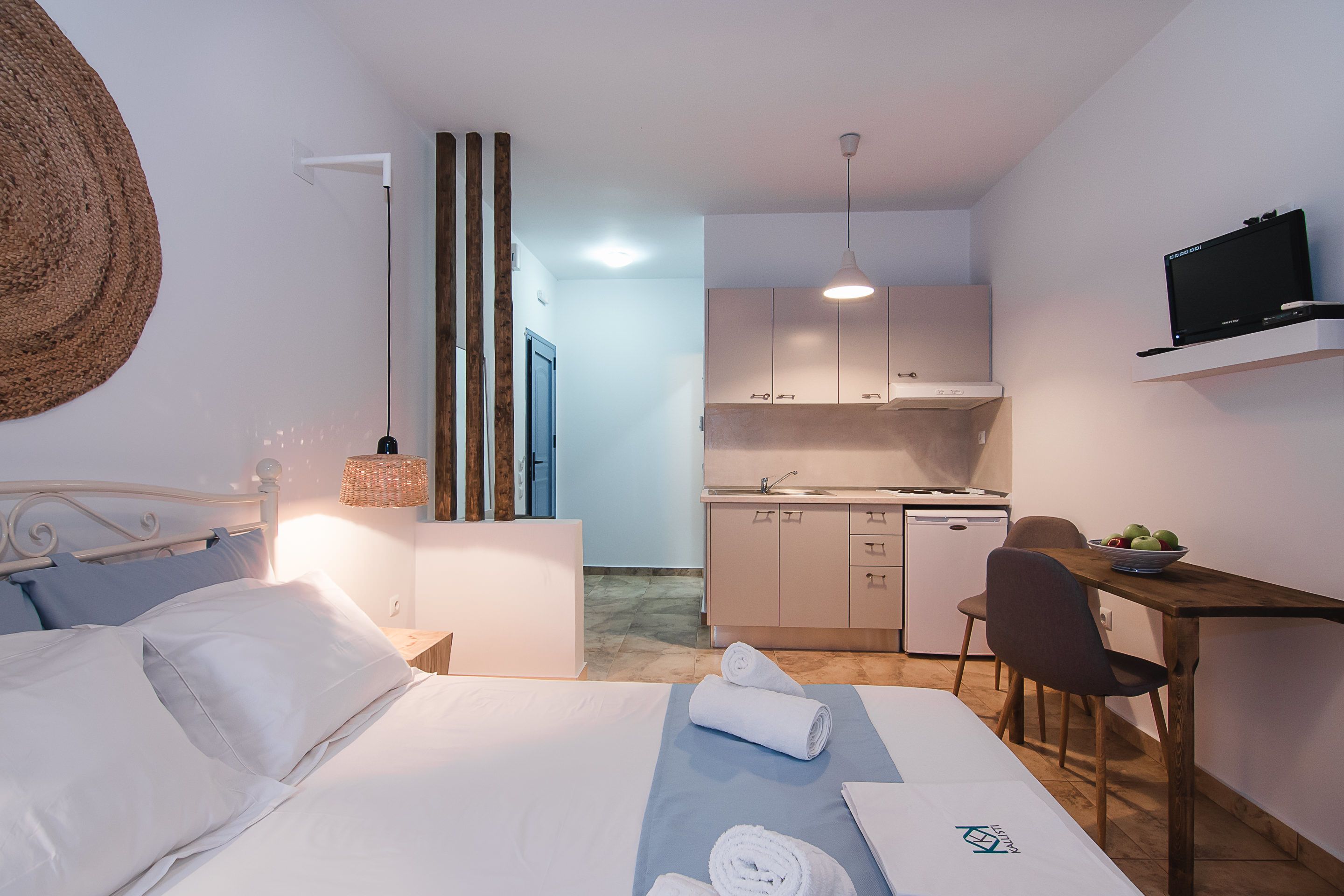 Quarto de hotel com cama, escrivaninha, cadeira, televisão e cozinha compacta.