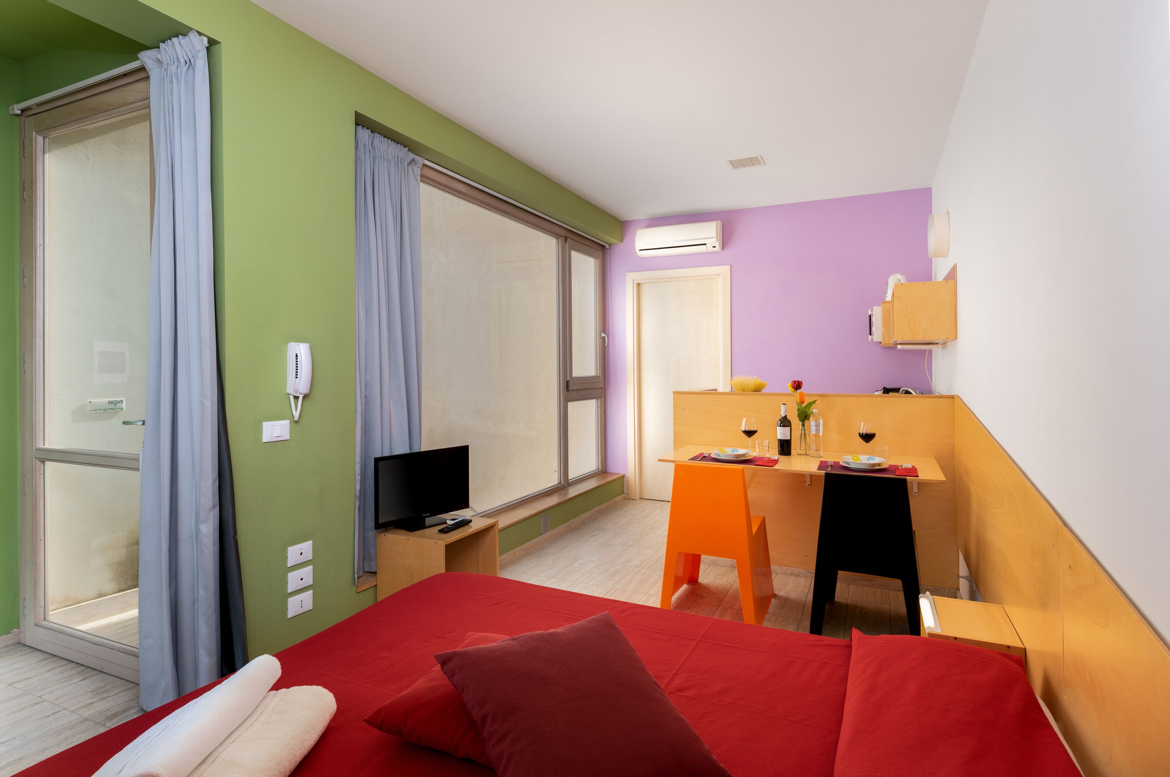 Quarto de hotel com cama vermelha, escrivaninha com cadeira, televisão e porta de correr.