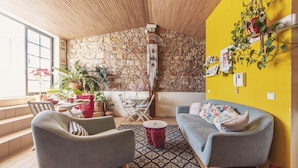 Uma sala de estar aconchegante com uma parede de pedra, teto de madeira e uma parede de destaque amarelo vibrante. Há um sofá, uma poltrona, uma mesinha e várias plantas em vasos.