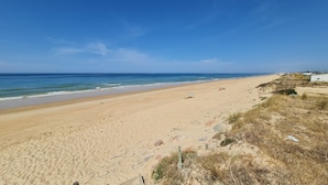 Uma extensa praia de areia com ondas suaves, céu claro e vegetação rala à direita.