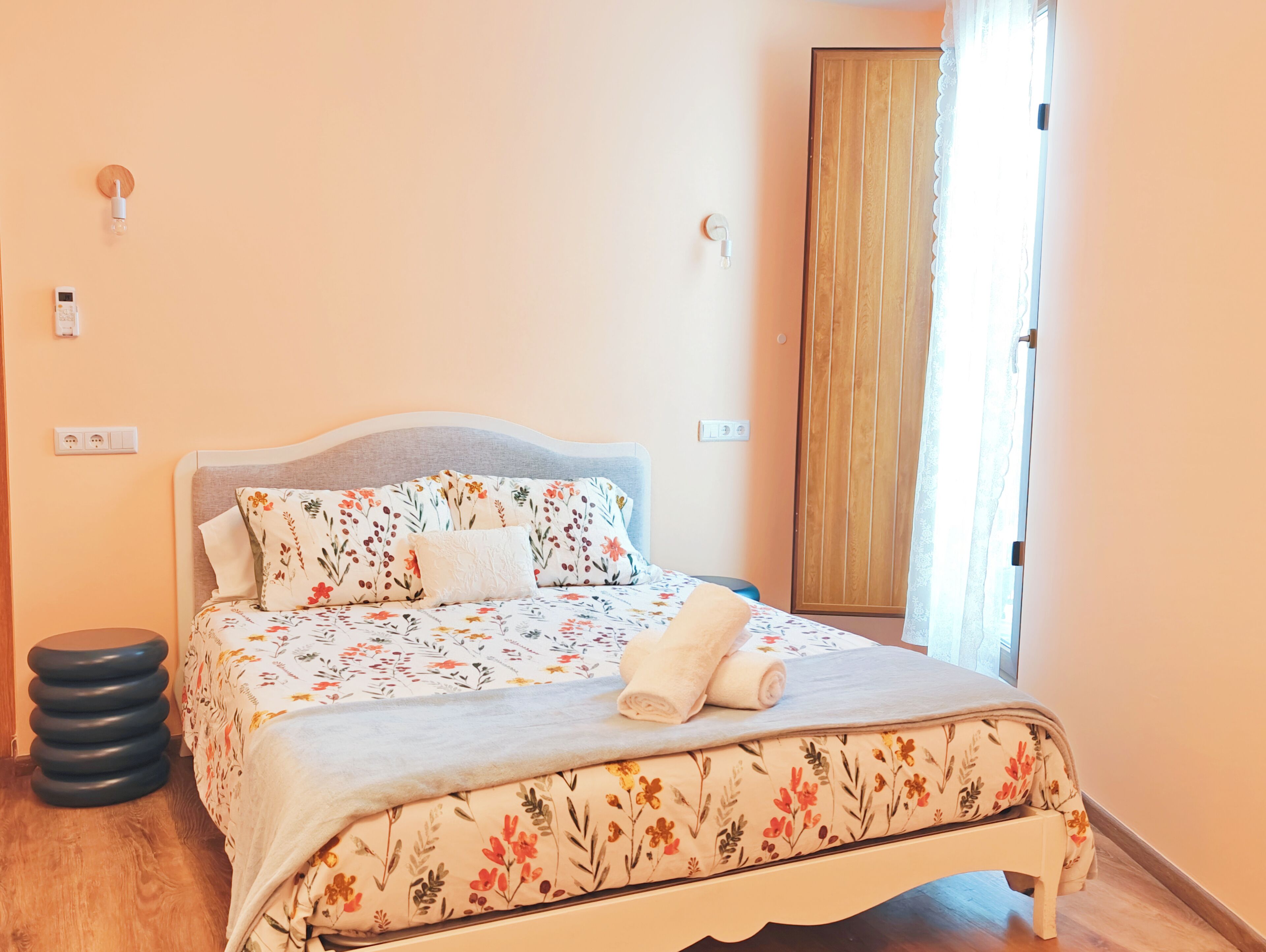 Cama bem arrumada com roupa de cama florida, mesa de cabeceira e porta de madeira em um quarto com paredes cor de pêssego.