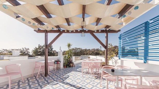 Um pátio externo com piso estampado geométrico, cadeiras e mesas cor-de-rosa, uma pérgola com luzes de corda e uma parede listrada em azul e branco.
