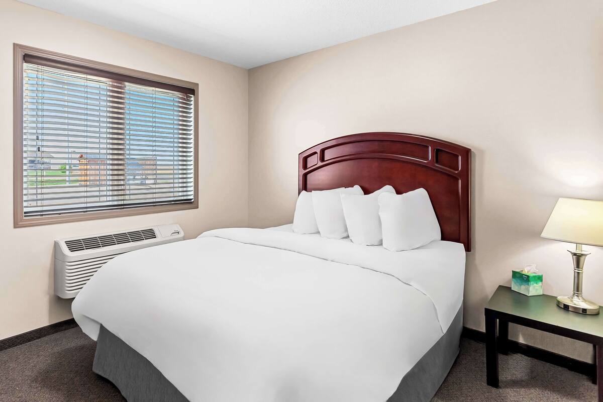 Quarto de hotel com uma cama grande, uma mesa de cabeceira com abajur e uma janela com persianas.