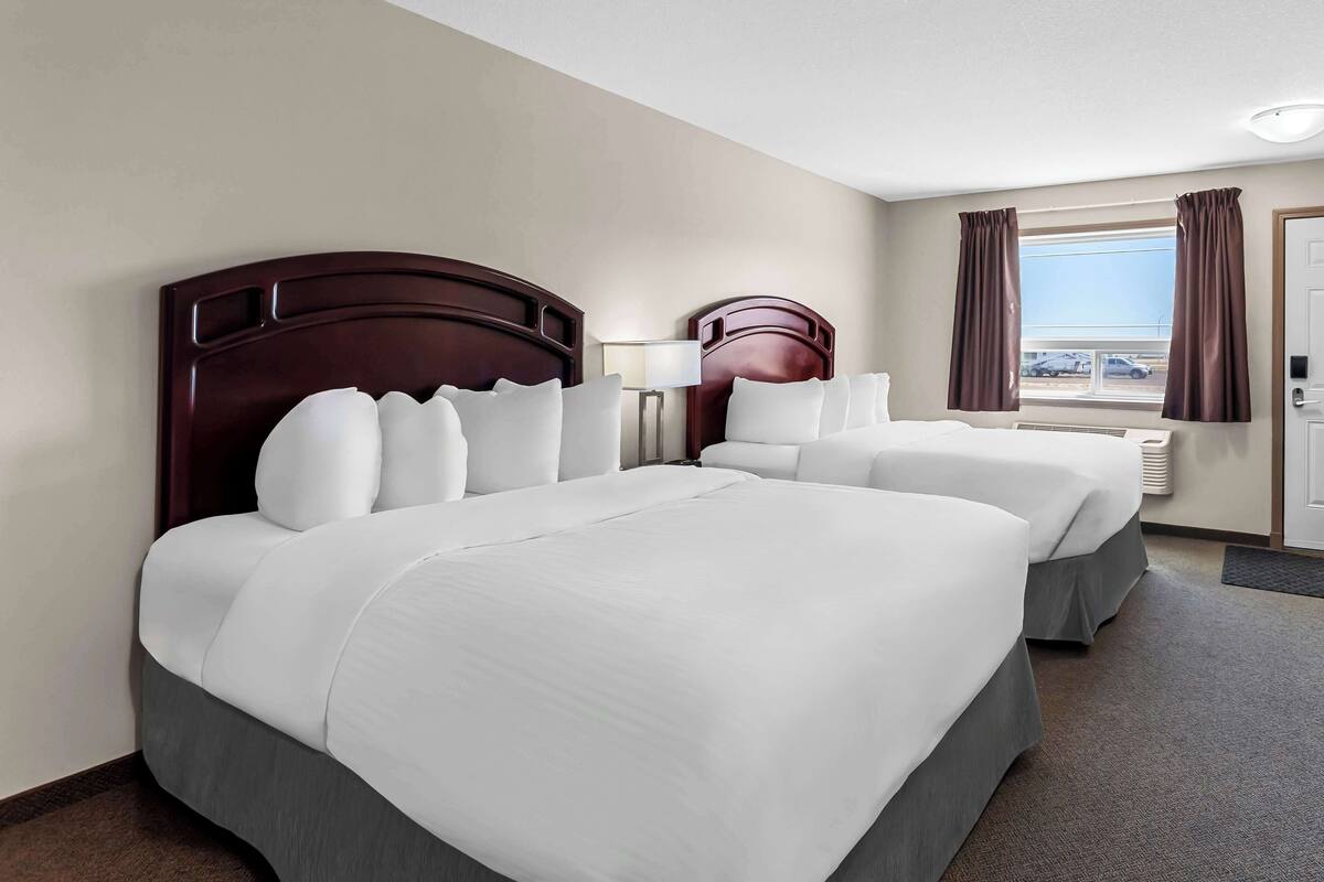 Quarto de hotel com duas camas, uma janela grande com cortinas, um abajur e uma porta.