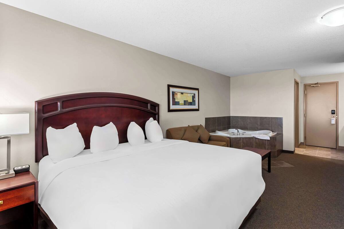 Quarto de hotel com uma cama grande, mesa de cabeceira, cadeira, mesinha e um quadro na parede, além de banheiro com banheira e pia.