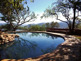 Piscina com borda de pedra, rodeada por árvores e vista para montanhas.
