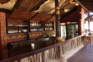 Uma área de bar com prateleiras de madeira repletas de garrafas e copos, um refrigerador de Coca-Cola e um aviso sobre programa de recompensas.
