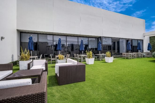 Uma área de estar moderna ao ar livre com mobiliário de vime branco e escuro, plantas em vasos e guarda-sóis azuis.