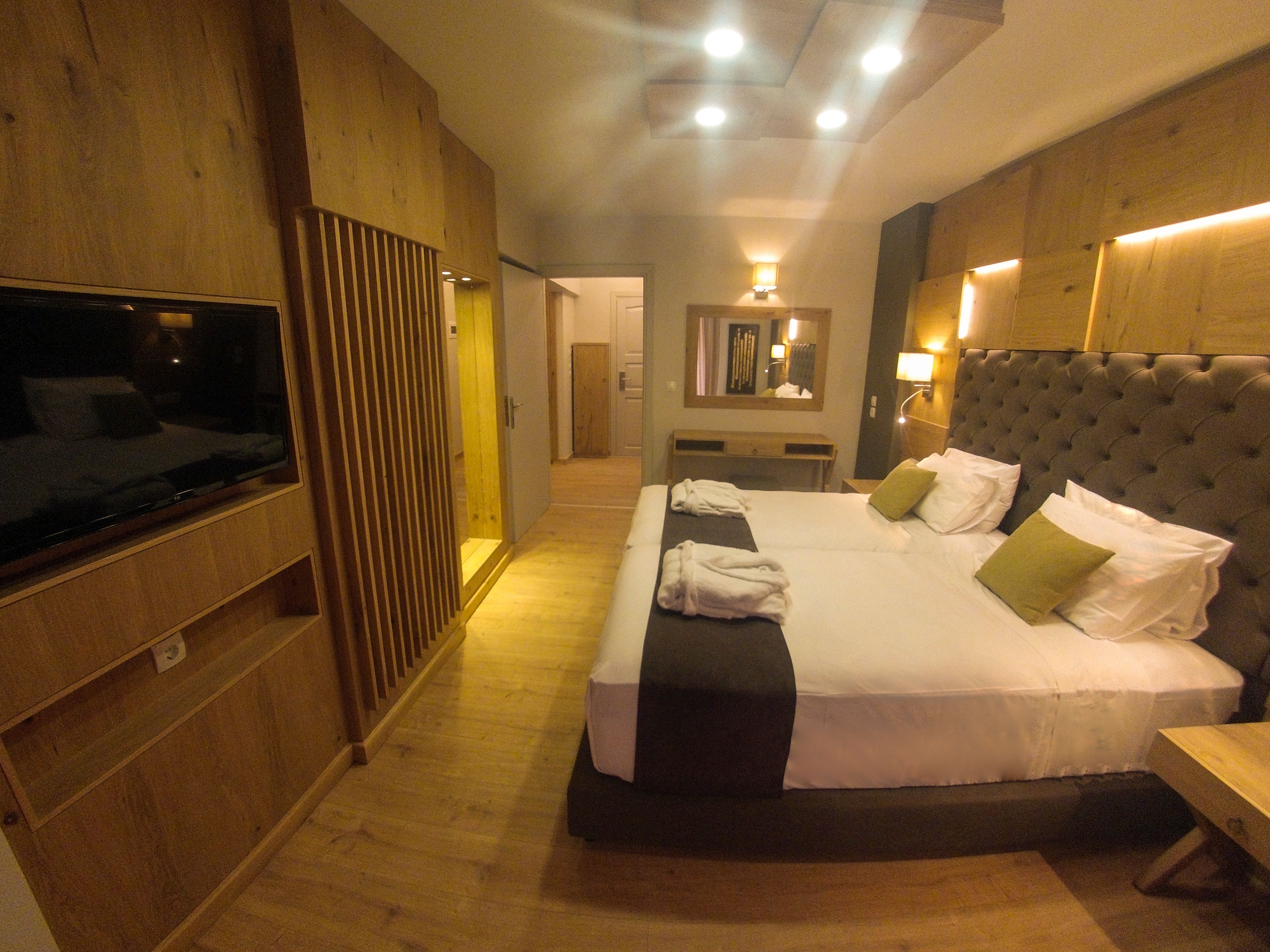 Quarto de hotel com uma cama grande, televisão na parede, guarda-roupa de madeira e espelho sobre a cama.