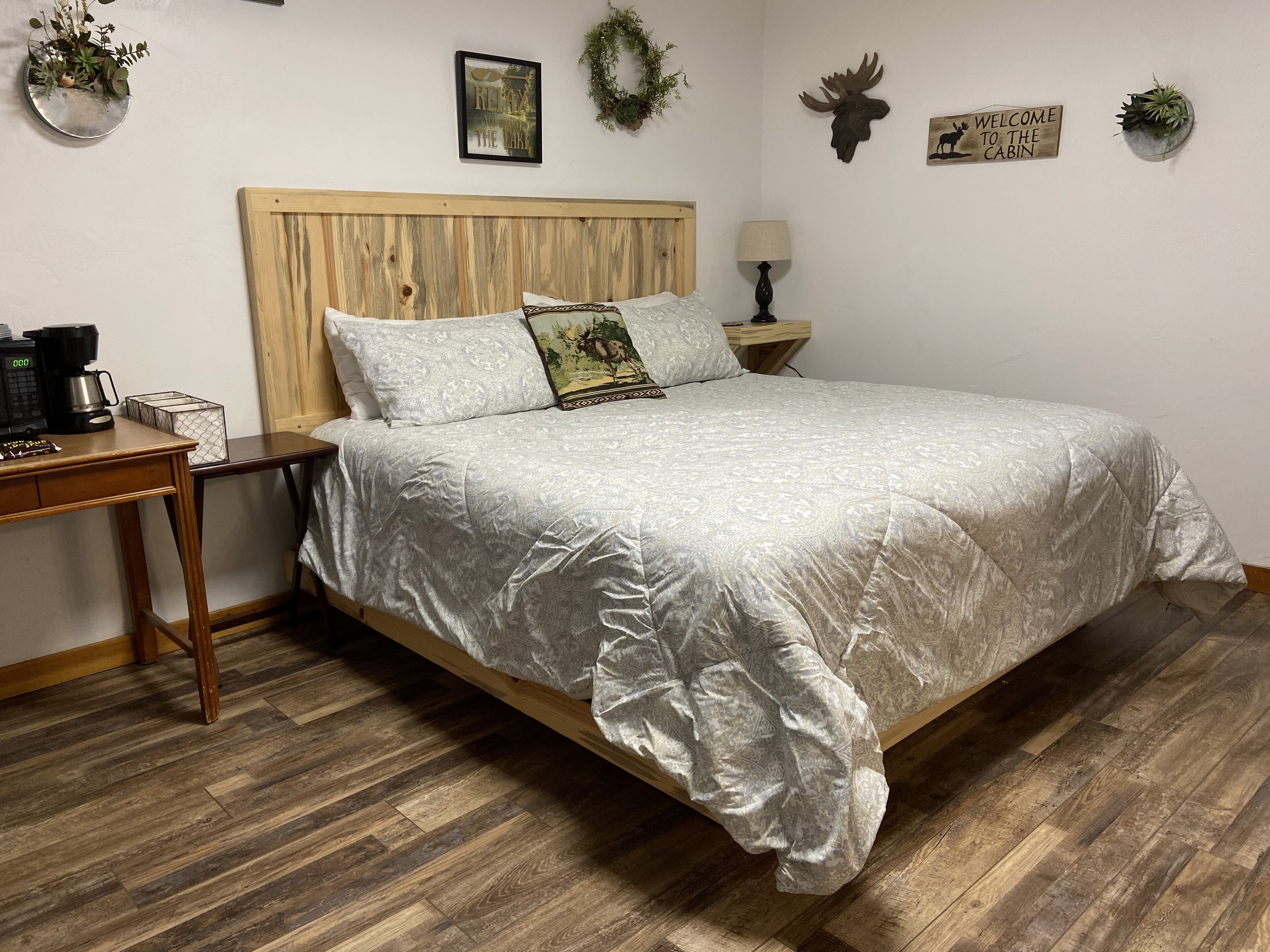 Quarto com cabeceira de madeira, cama com roupa de cama estampada, mesa de cabeceira com cafeteira, luminária e decoração de parede incluindo uma placa de boas-vindas e uma cabeça de alce.