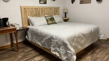 Quarto com cabeceira de madeira, cama com roupa de cama estampada, mesa de cabeceira com cafeteira, luminária e decoração de parede incluindo uma placa de boas-vindas e uma cabeça de alce.