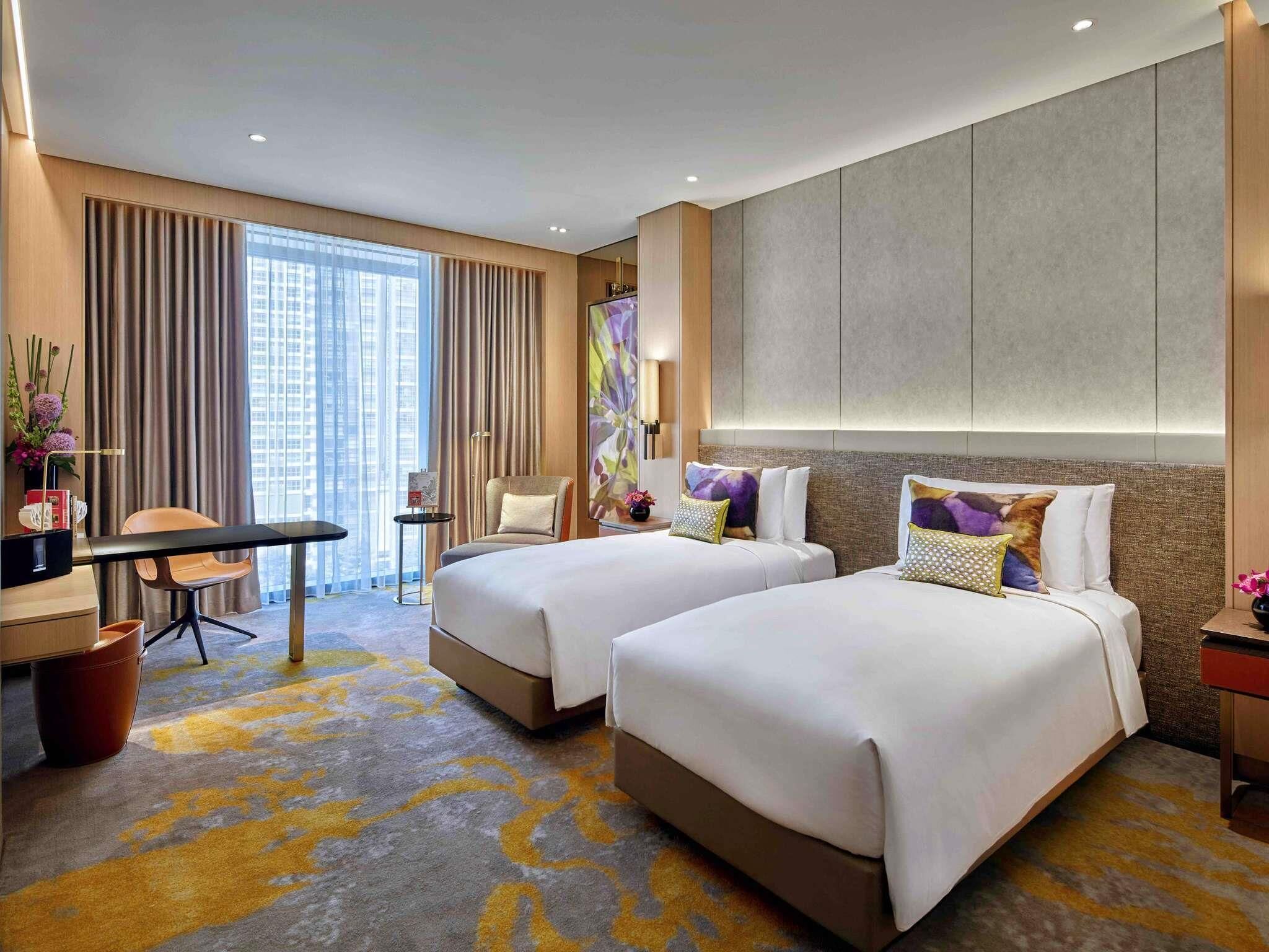 Quarto de hotel com duas camas, uma escrivaninha, uma cadeira e uma janela grande com cortinas.