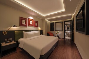 Quarto de hotel com uma cama grande, uma mesa de cabeceira, uma escrivaninha, uma cadeira e um banheiro visível por uma porta aberta.