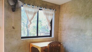 Quarto com escrivaninha e cadeira de madeira, janela com cortinas e ventoinha fixada na parede.
