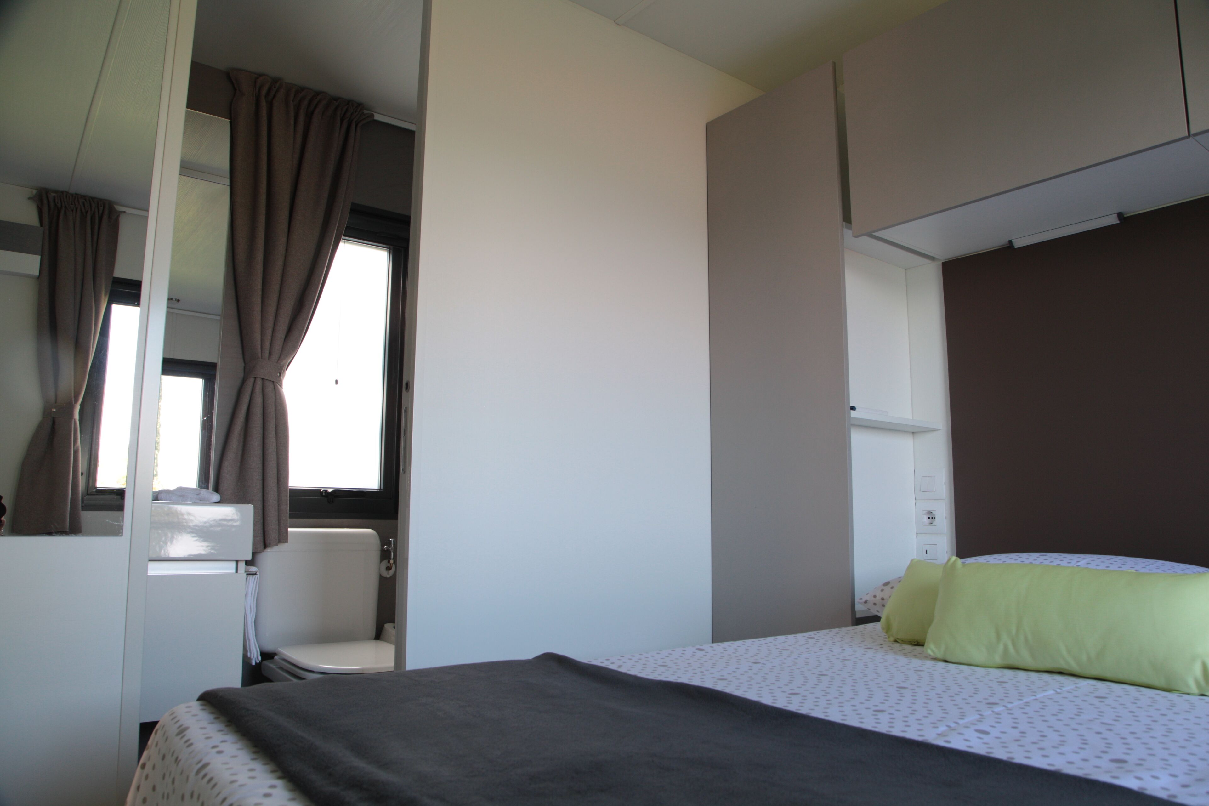 Um quarto com cama, janela com cortinas, um guarda-roupa branco e uma mesinha.