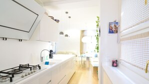 Uma cozinha moderna com armários brancos, fogão a gás e pia. É possível ver uma área de jantar com mesa e cadeiras através de uma porta aberta.