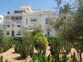 Um edifício branco com várias janelas e varandas, cercado por vegetação e carros estacionados.