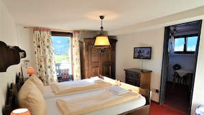 Quarto com cama de casal, guarda-roupa de madeira, escrivaninha com cadeira e televisão na parede.