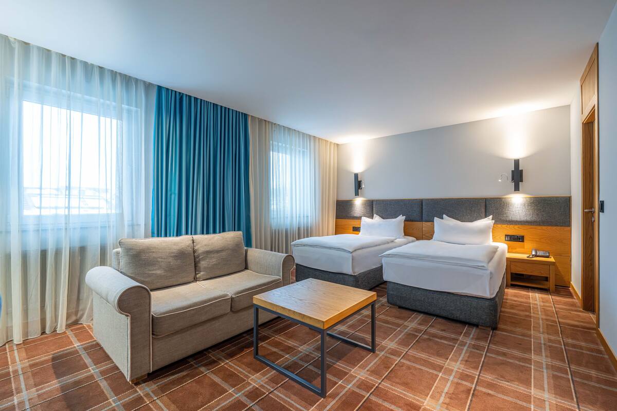 Quarto de hotel com sofá, duas camas de solteiro, uma mesinha e uma mesa de cabeceira com abajur.