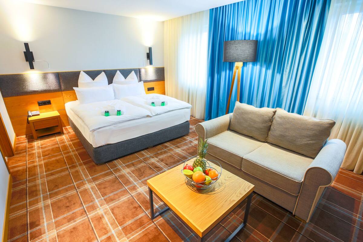 Quarto de hotel com cama, sofá, mesa de centro com frutas e mesa de cabeceira.