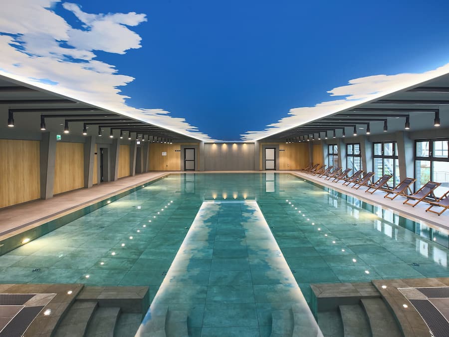 Uma grande piscina interior com vista clara para o céu através do teto, rodeada por espreguiçadeiras e um design arquitetônico moderno.
