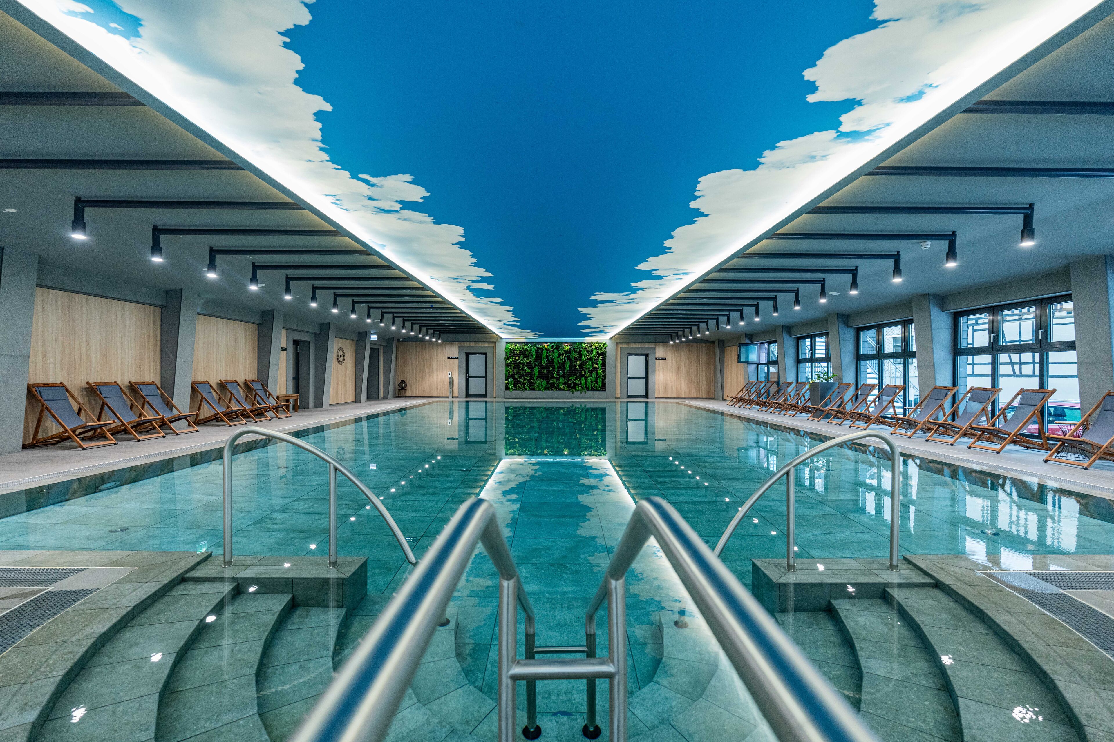 Piscina interior moderna com águas cristalinas, espreguiçadeiras e teto com design inspirado em nuvens.
