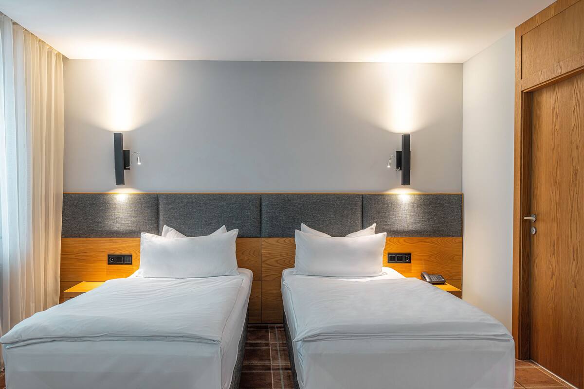 Quarto de hotel com duas camas de solteiro, cabeceira de madeira, mesa de cabeceira com telefone e luminária na parede.