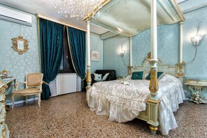 Uma cama de dossel com detalhes dourados ornamentais, um espaço de estar com uma poltrona e uma mesa pequena, e um espelho grande com moldura dourada.