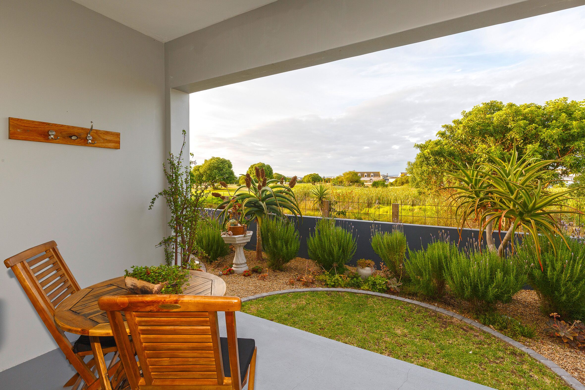 Um pátio com mesa e cadeiras de madeira, rodeado por um jardim com diversas plantas e vista para um campo.