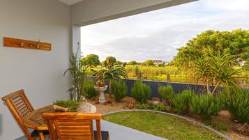 Um pátio com mesa e cadeiras de madeira, rodeado por um jardim com diversas plantas e vista para um campo.