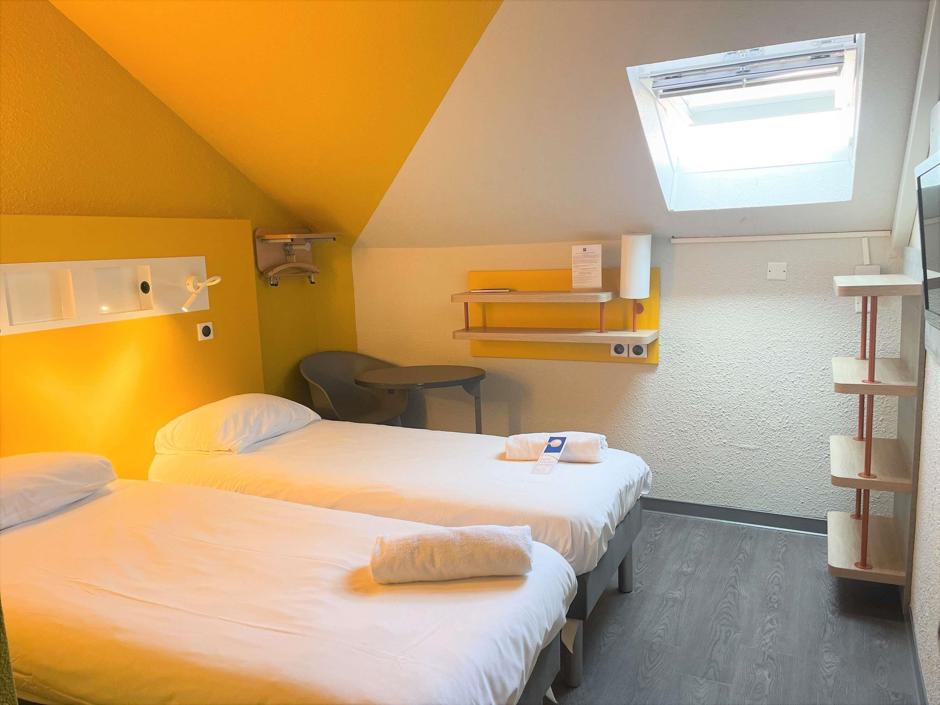 Quarto de hotel com duas camas, uma mesa pequena, uma cadeira, uma escrivaninha com prateleiras e um claraboia.