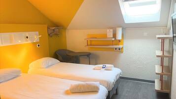 Quarto de hotel com duas camas, uma mesa pequena, uma cadeira, uma escrivaninha com prateleiras e um claraboia.