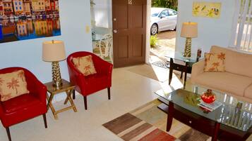 Uma sala de estar com uma poltrona vermelha, um sofá bege, uma mesa de centro de vidro e uma porta que dá para o exterior.