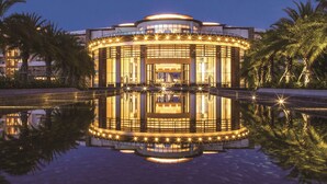 Um edifício moderno com design circular, uma piscina refletora à frente e palmeiras ao redor.