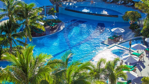 Uma grande piscina ao ar livre com várias pistas de natação, rodeada por espreguiçadeiras e guarda-sóis, e palmeiras.