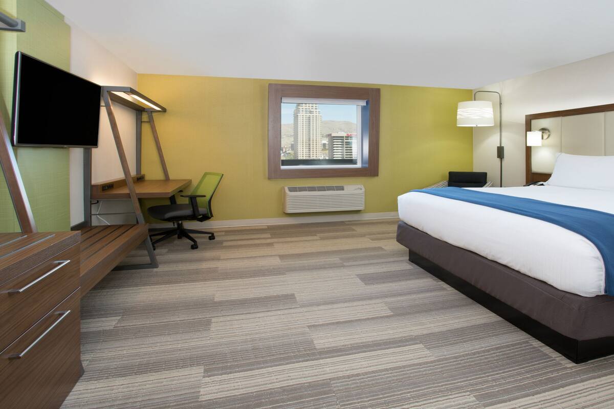 Quarto de hotel com cama, escrivaninha, cadeira, televisão, janela e abajur.