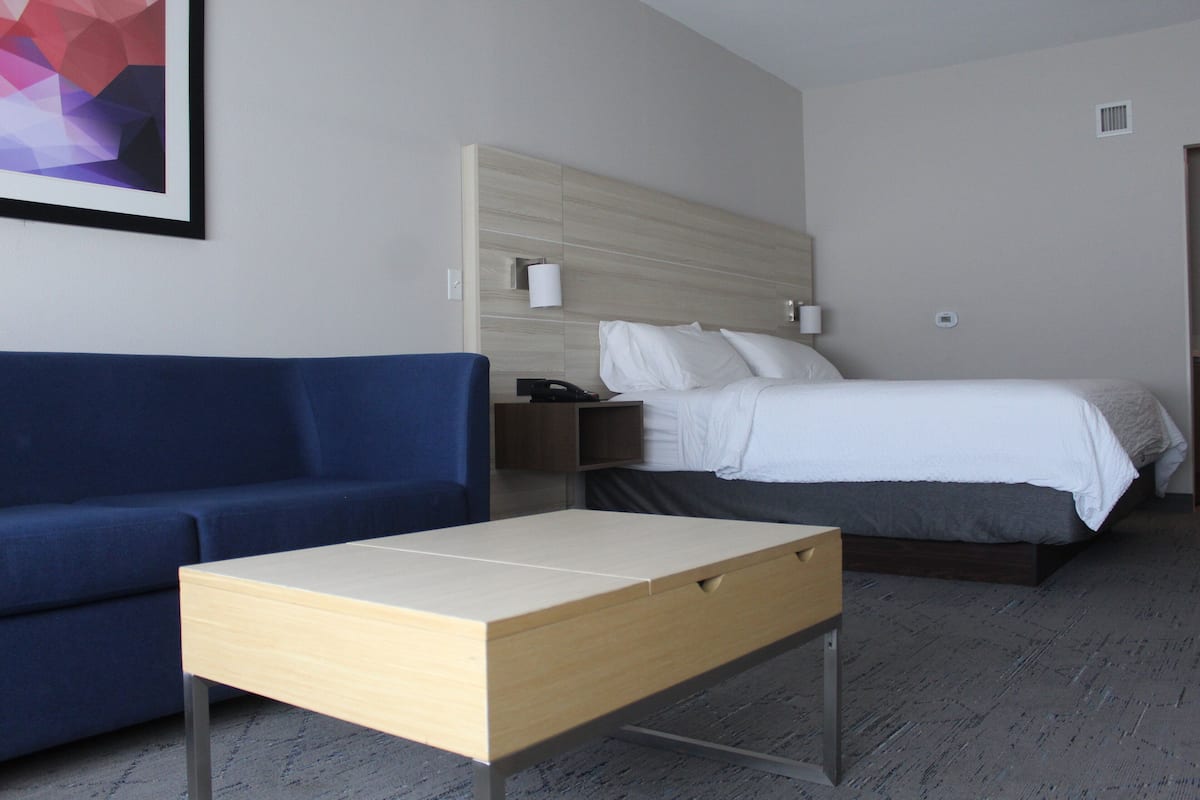 Quarto de hotel moderno com cama, sofá azul, mesa de cabeceira de madeira e luz acoplada à parede.