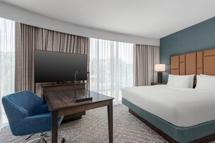 Quarto de hotel com cama, secretária com televisão de tela plana, cadeira, abajur e vista do exterior através de amplas janelas.