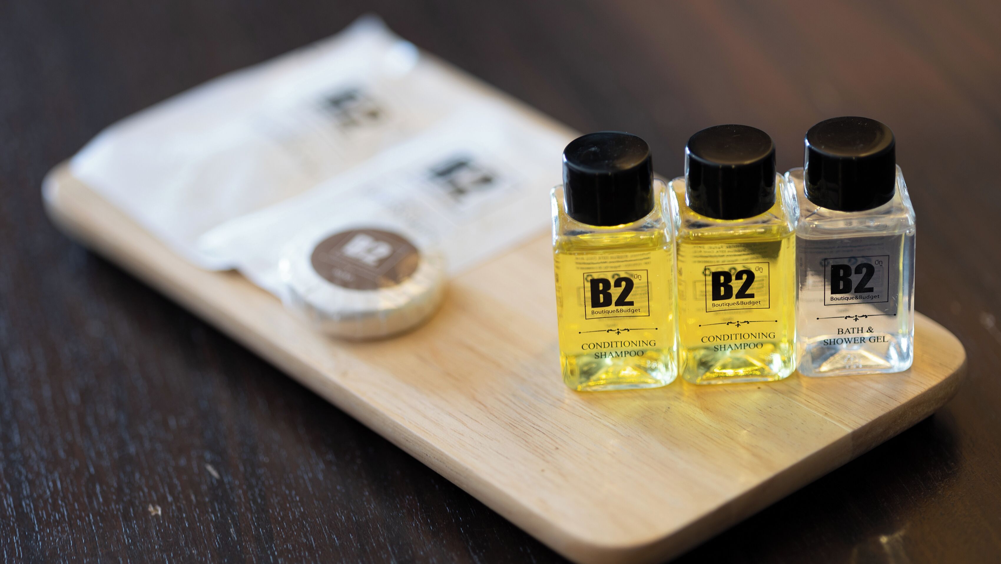 Três frascos de shampoo condicionante B2, gel de banho e chuveiro B2, e uma nécessaire branca sobre uma bandeja de madeira.