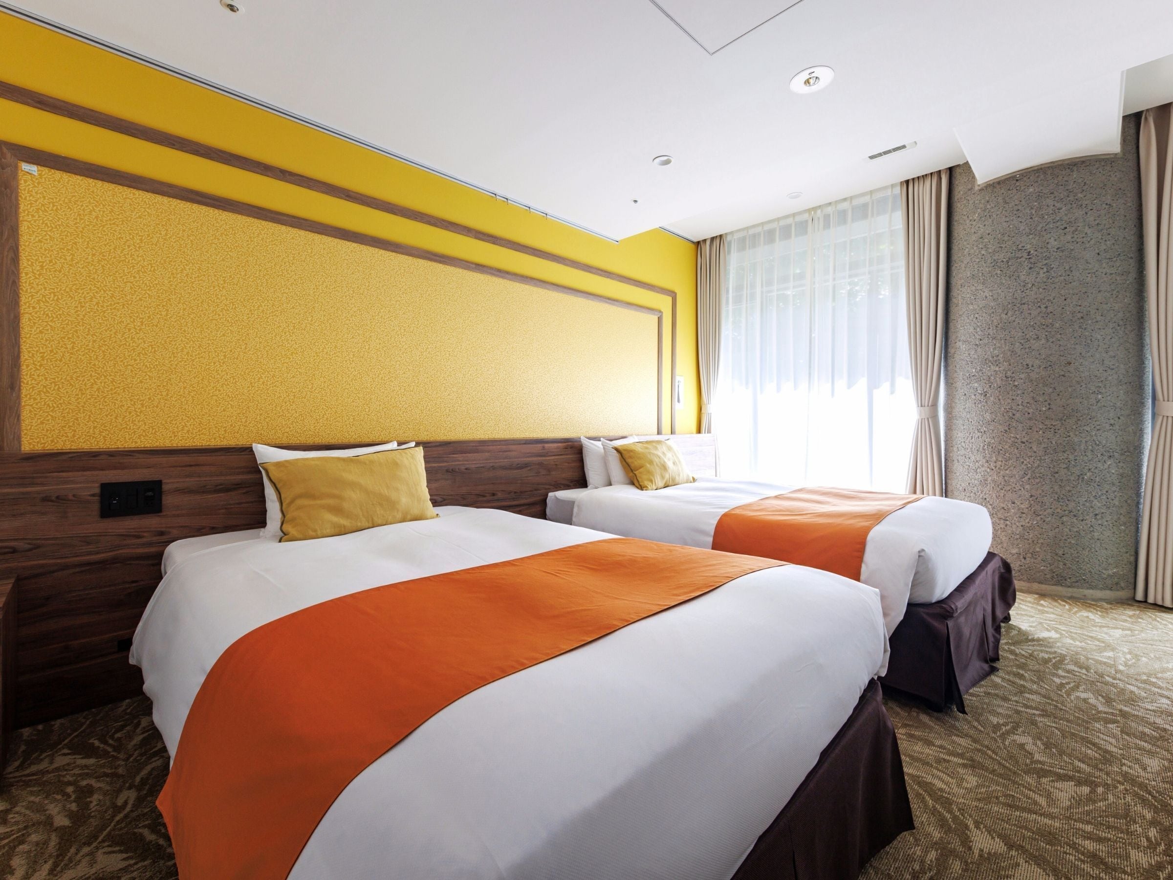 Quarto de hotel com duas camas, cabeceira de madeira, parede amarela e uma grande janela com cortinas.