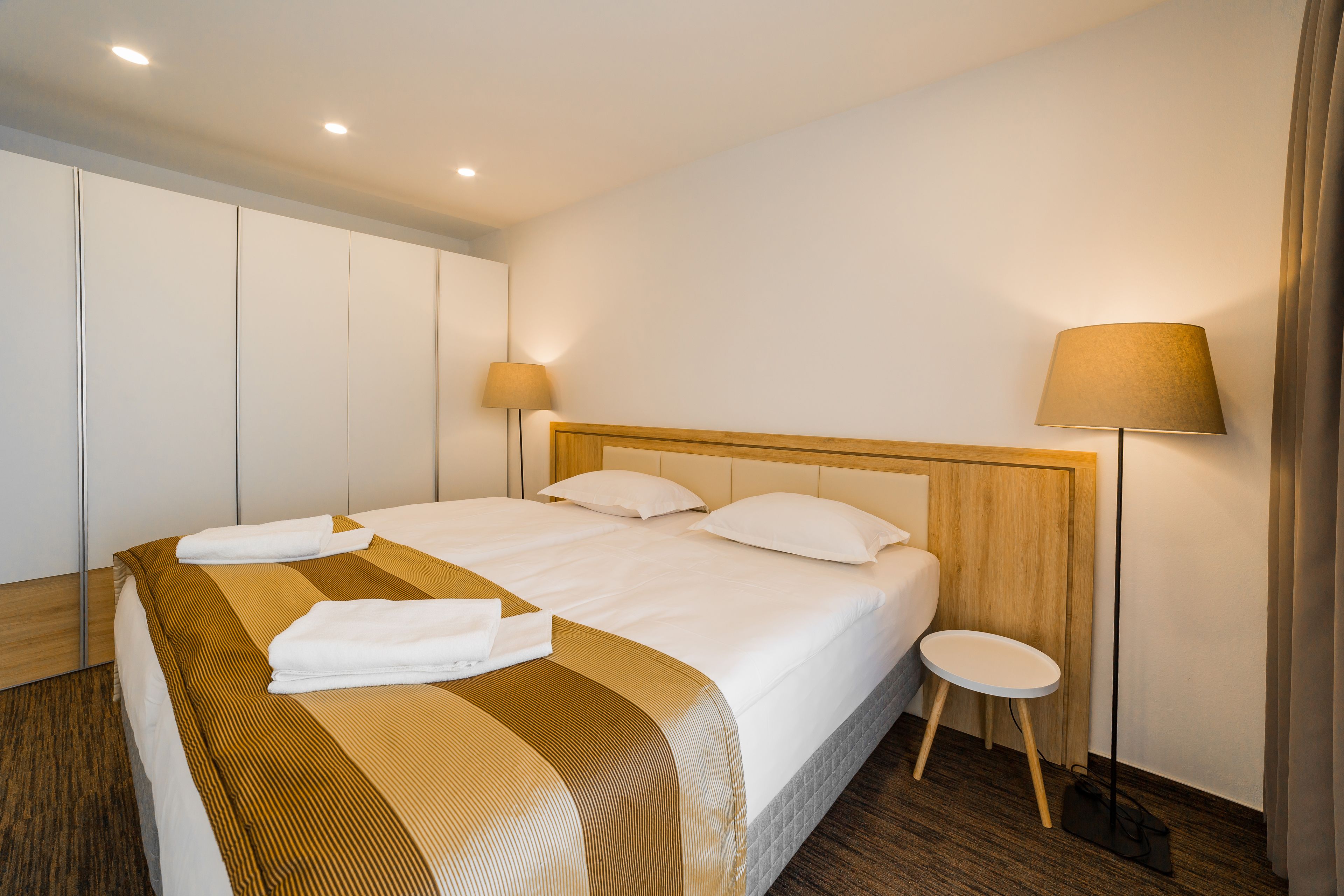 Quarto de hotel com uma cama grande, duas luminárias de cabeceira, um painel de cama de madeira e uma mesa redonda pequena.