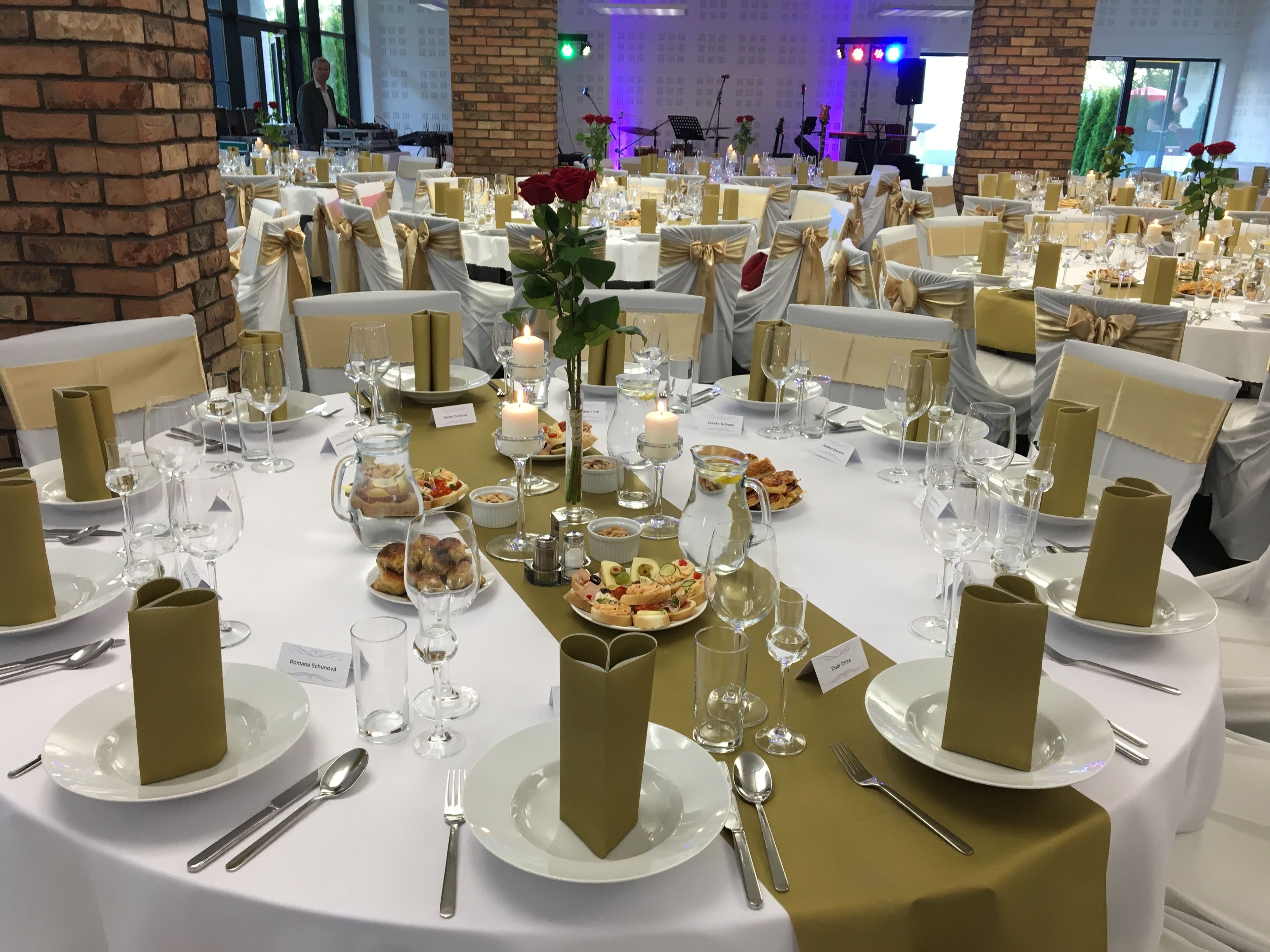 Uma sala de banquetes com mesas redondas preparadas para um evento formal, com toalhas brancas, fitas douradas e centros de mesa com velas.