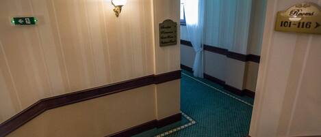 Corredor de hotel com paredes listradas, carpete com padrão verde e placas de sinalização indicando comodidades e números de quartos.