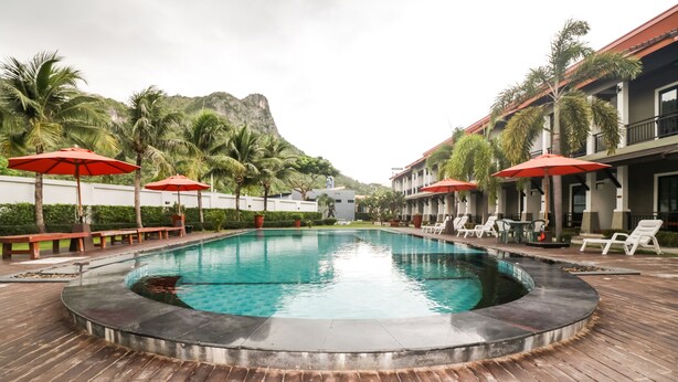 Área de piscina com deck de madeira, guarda-sóis vermelhos, espreguiçadeiras e montanhas ao fundo.