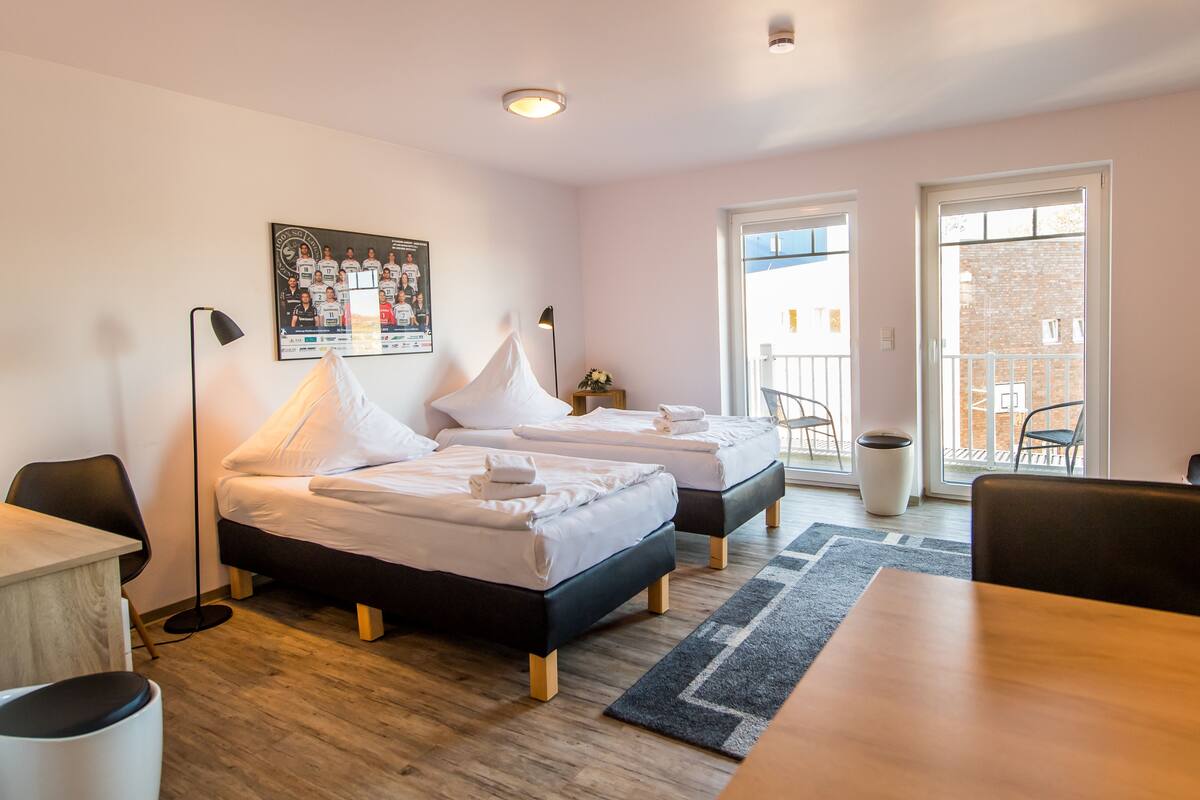 Quarto de hotel com duas camas, uma escrivaninha, uma cadeira, um abajur e vista para a varanda.