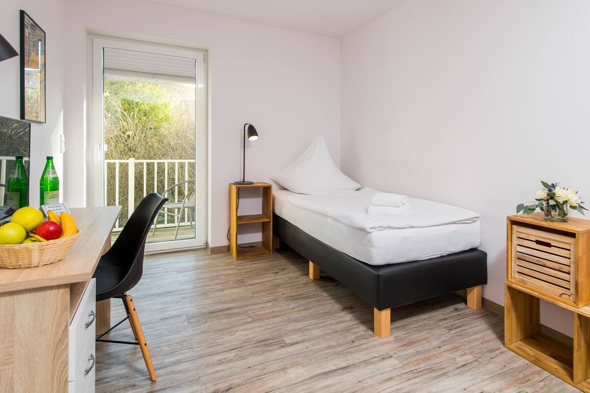 Quarto com cama, escrivaninha e cadeira, mesinha de cabeceira e criado-mudo. Há uma janela com vista para o verde lá fora.